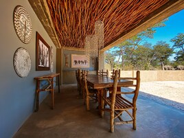 Uma sala de jantar com uma longa mesa de madeira e cadeiras, rodeada por paredes decoradas com diversas obras de arte, sob um telhado de palha.