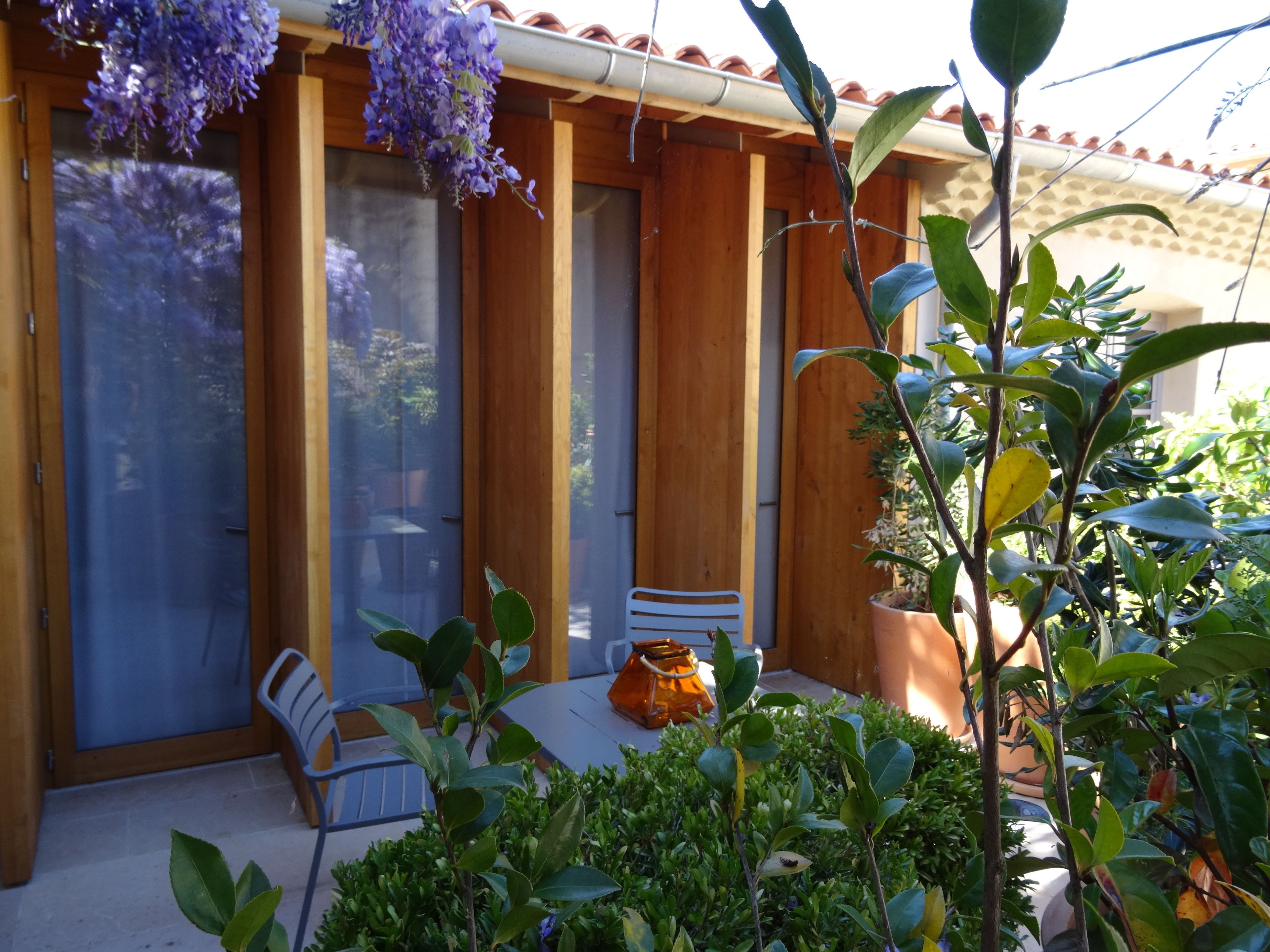 Uma pérgola de madeira com flores roxas, uma cadeira branca e uma mesa pequena com uma garrafa.