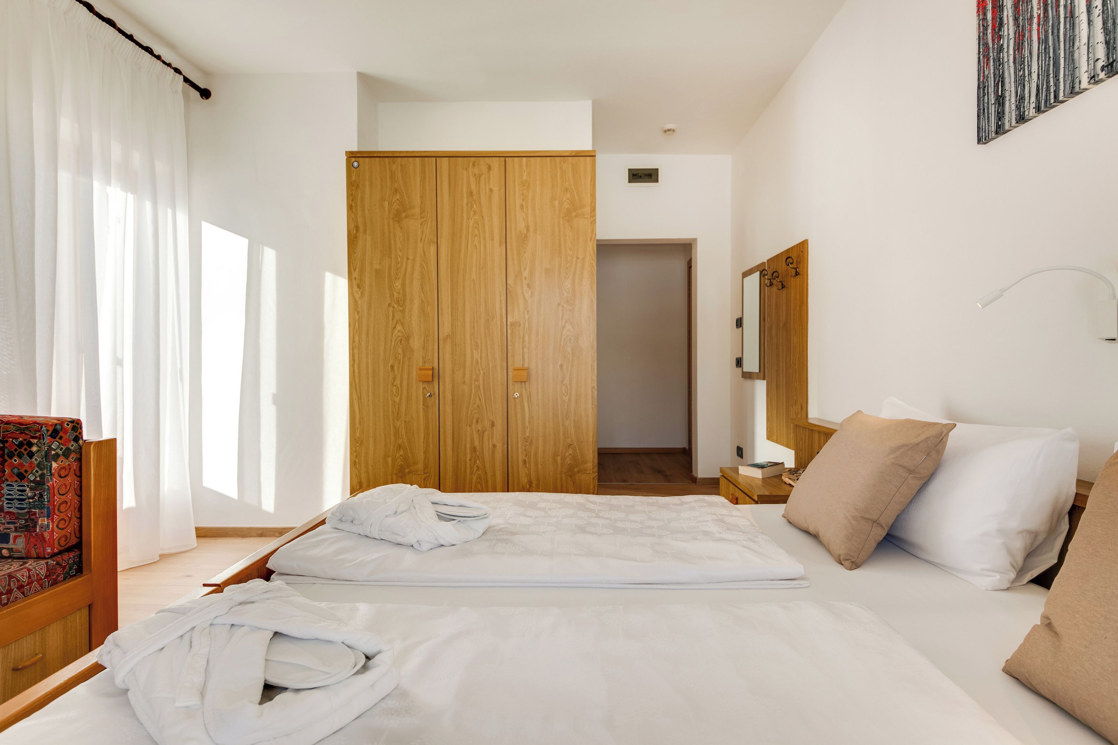 Quarto de hotel com cama, guarda-roupa de madeira, espelho e janela com cortinas.