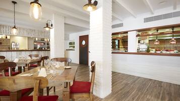 Um restaurante com mesas e cadeiras de madeira, área de bar e cozinha visível através de um separador de vidro.
