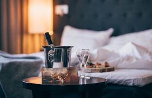Uma cama com roupa branca, uma mesa de cabeceira com uma garrafa de champanhe, um copo e um menu.