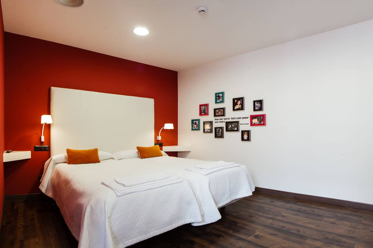 Quarto de hotel com uma cama grande, duas luminárias de cabeceira, uma parede de destaque vermelha e uma parede com molduras de fotos.