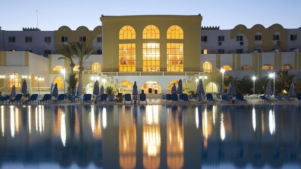 Um hotel com uma piscina grande refletindo luzes, uma área externa coberta com espreguiçadeiras e um edifício com janelas em arco.