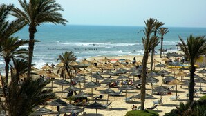 Uma praia com vários guarda-sóis de palha e palmeiras, oferecendo sombra para quem aproveita a praia.