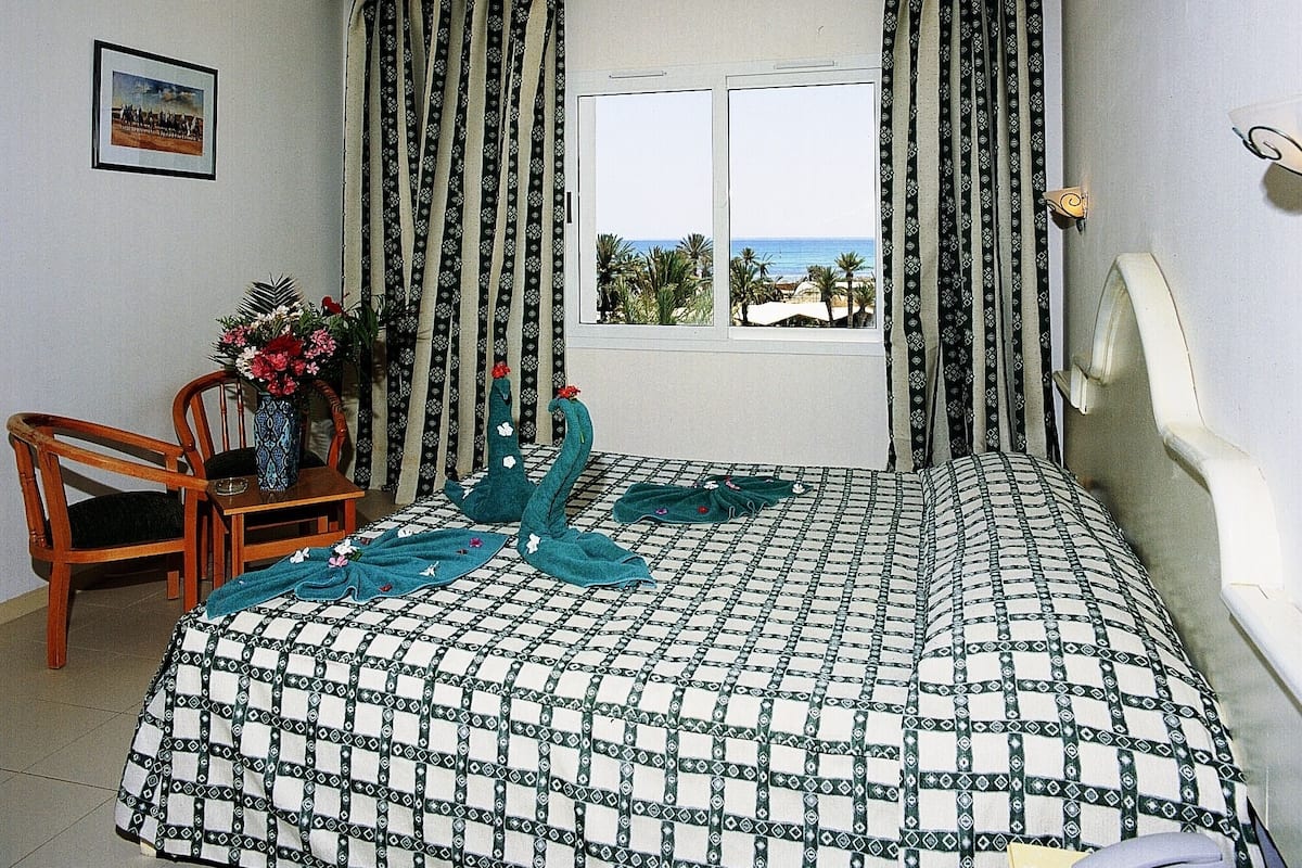 Quarto com cama, cadeira, mesa com um vaso de flores e cortinas.
