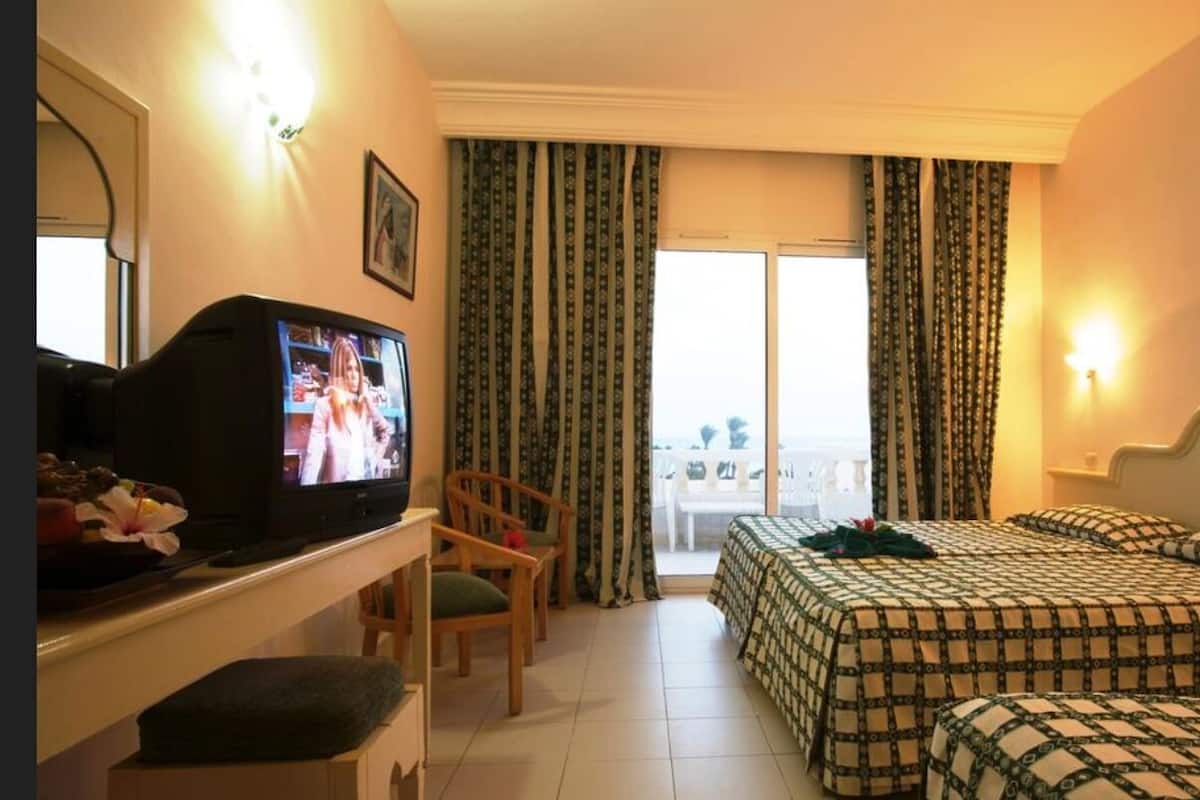 Quarto de hotel com televisão, escrivaninha, duas camas, uma cadeira e varanda com cortinas.