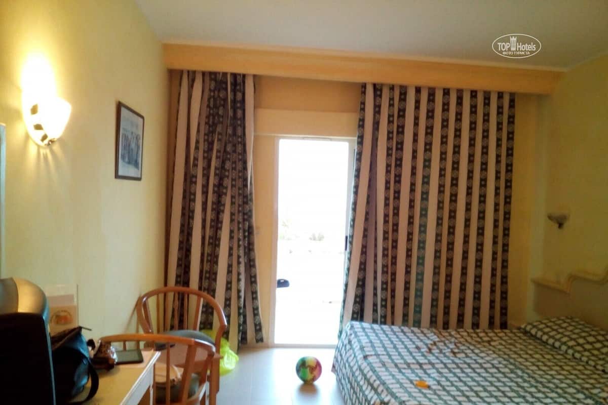 Quarto de hotel com cama, escrivaninha com cadeira, janela com cortinas e porta.
