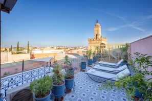 Terraço no telhado com espreguiçadeiras, plantas em vasos e vista para uma torre histórica.