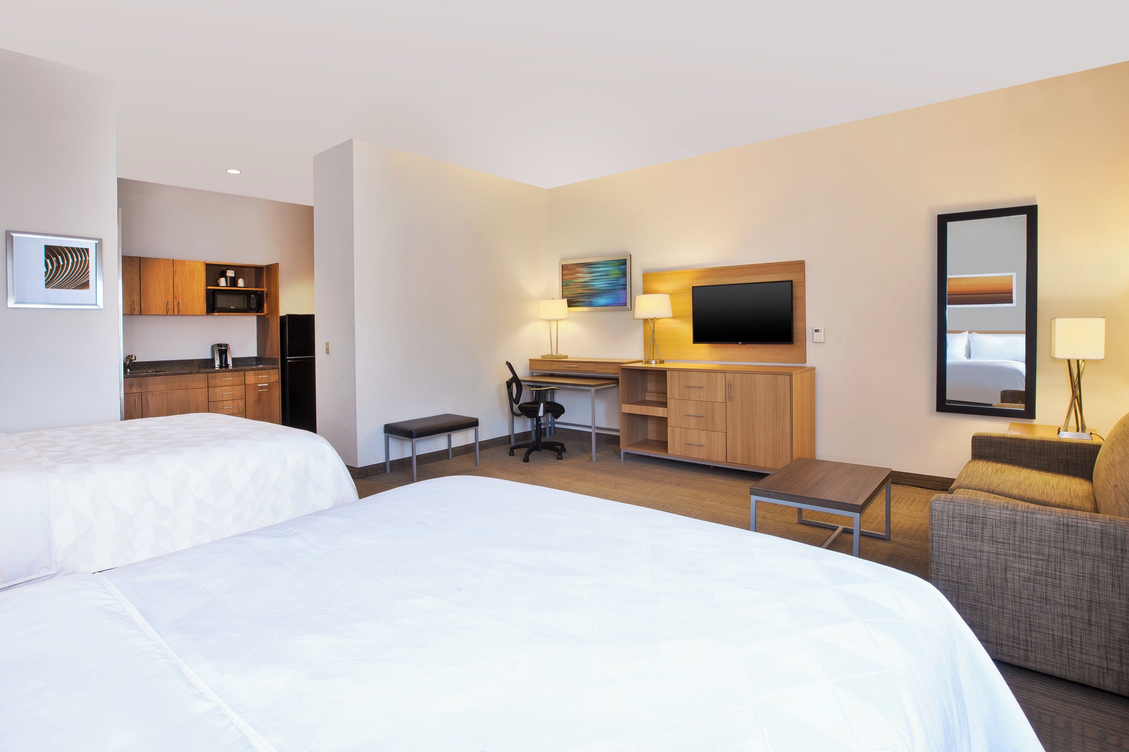 Quarto de hotel com duas camas, uma mesa, uma cadeira, televisão e cozinha compacta.
