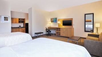 Quarto de hotel com duas camas, uma mesa, uma cadeira, televisão e cozinha compacta.