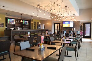 Um restaurante com várias mesas e cadeiras, área de bar com televisão e um jogo de esportes exibido em tela.