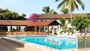 Piscina com espreguiçadeiras e guarda-sóis, cercada por palmeiras e um edifício com telhado em azulejos.