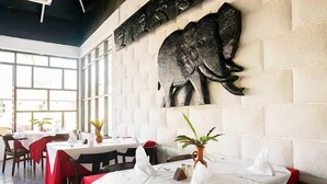 Restaurante com paredes brancas, uma grande escultura de elefante e mesas com toalhas brancas e guardanapos vermelhos.