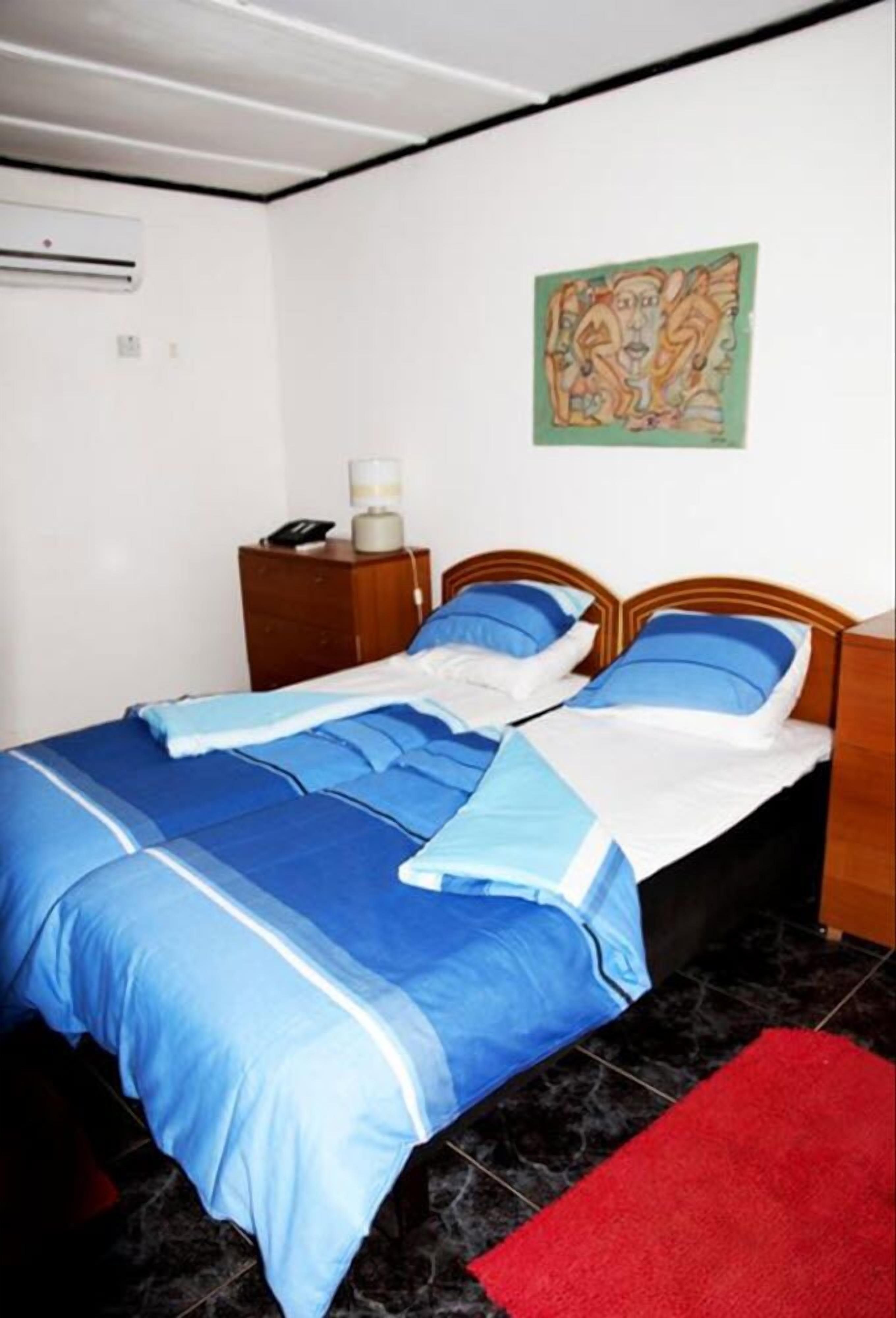 Quarto com duas camas, um tapete vermelho, um quadro na parede e uma mesinha de cabeceira com abajur e telefone.
