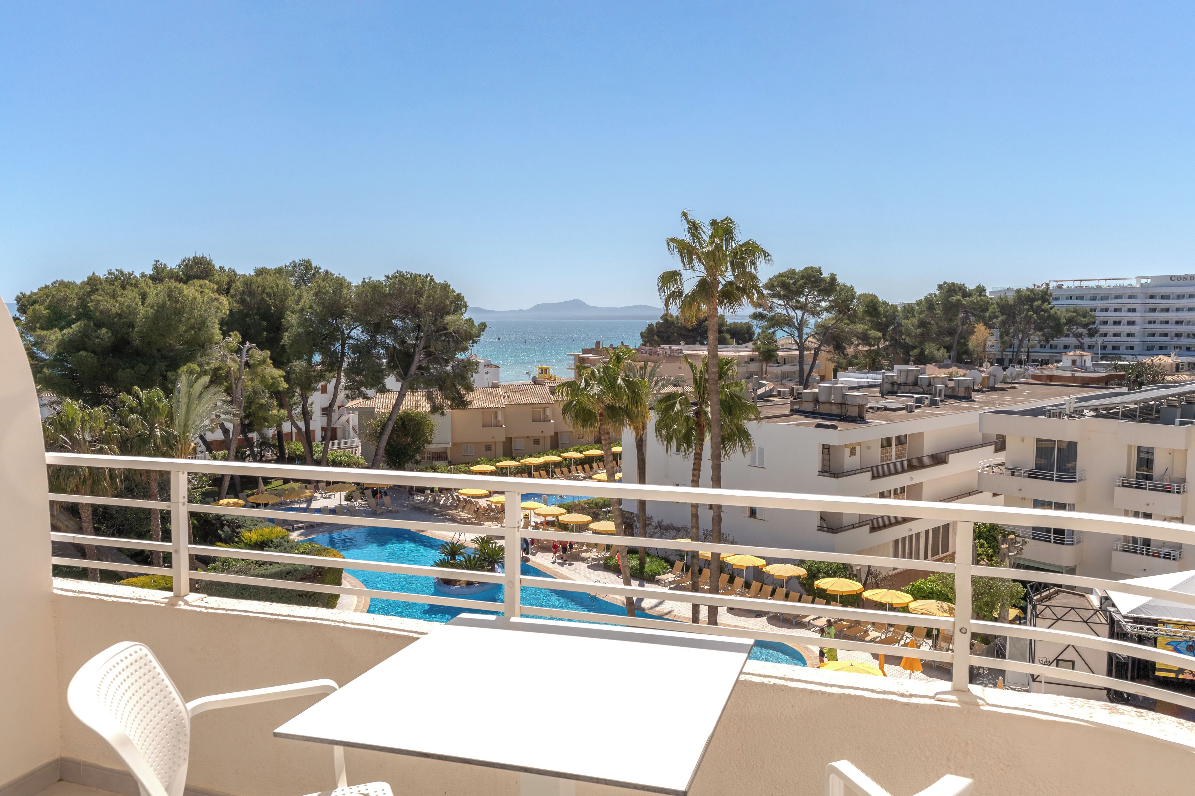 Varanda com piscina, espreguiçadeiras e guarda-sóis, com vista para uma área costeira com edifícios e árvores.