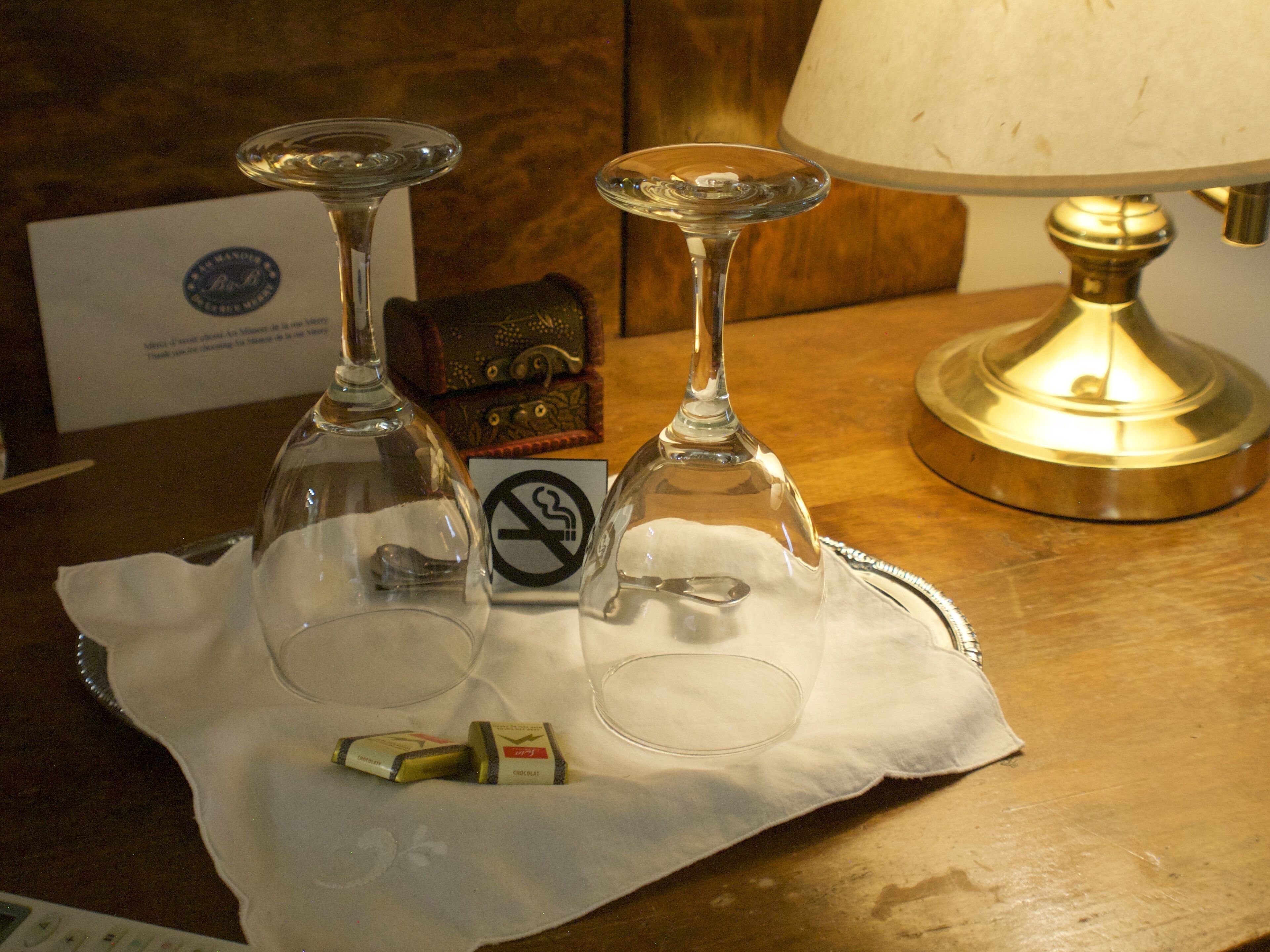 Duas taças de vinho vazias sobre uma mesa, com um abajur aceso e um aviso de proibido fumar ao fundo.