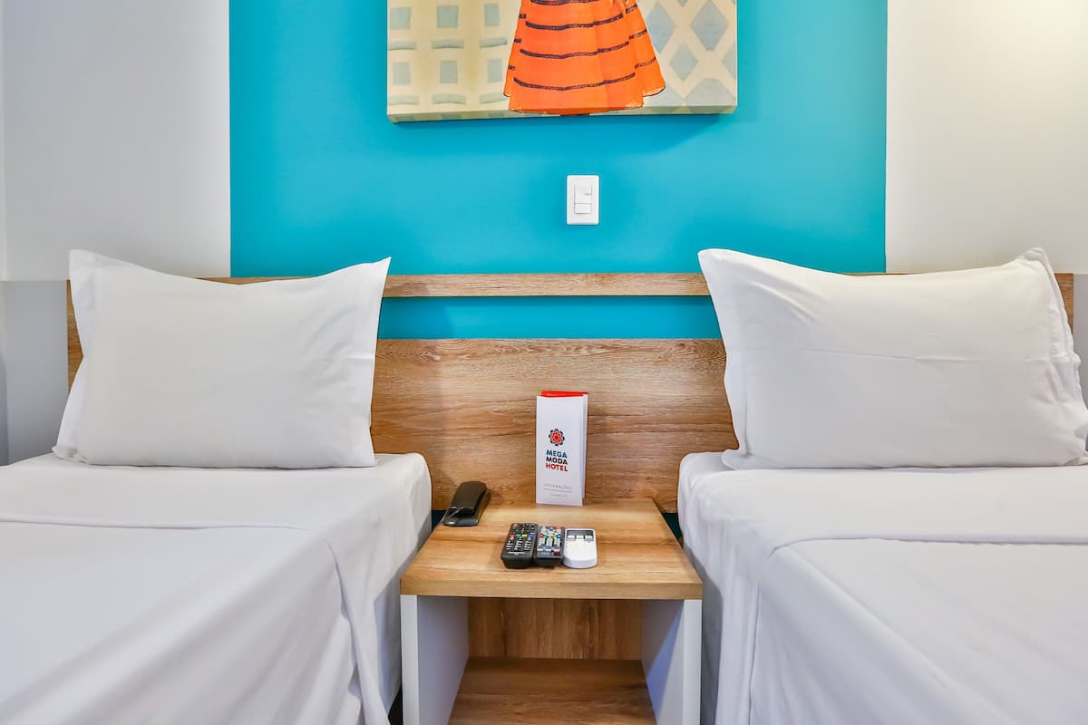 Quarto de hotel com duas camas de solteiro, uma mesinha de cabeceira de madeira, um controle remoto, um telefone e uma placa para o Mega Moda Hotel.