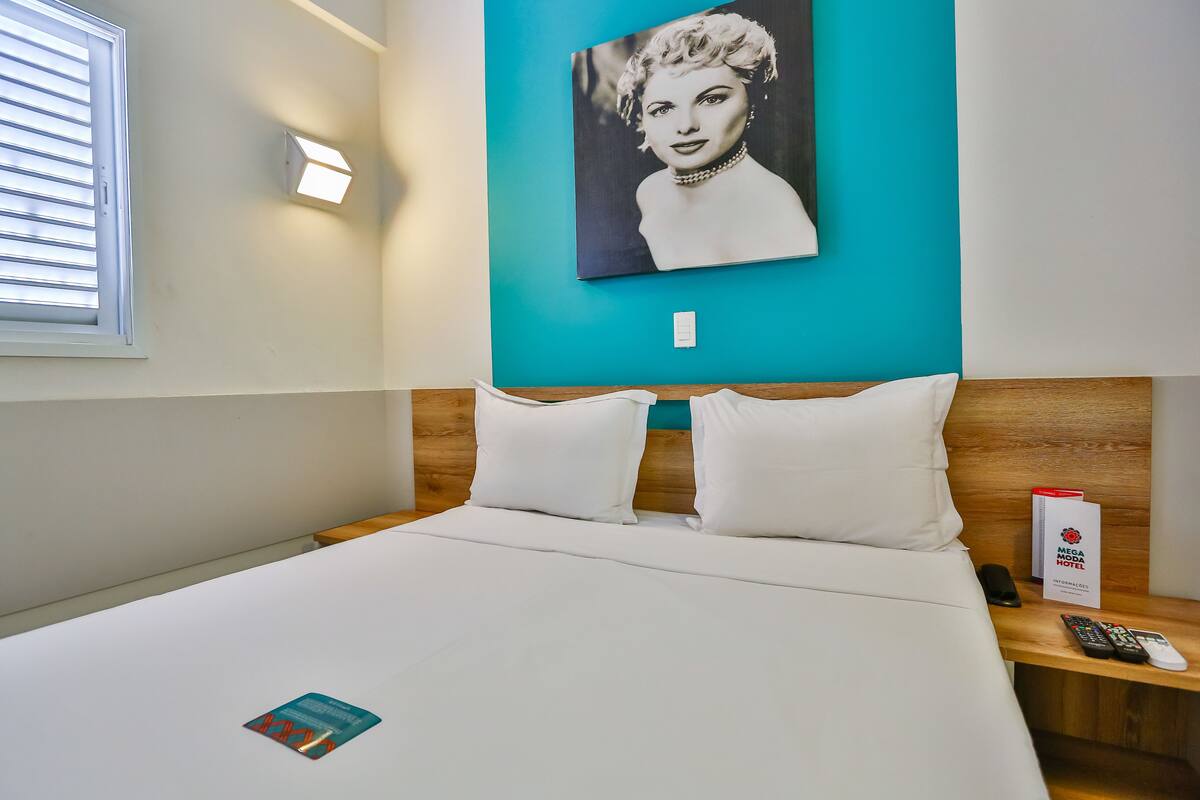 Quarto de hotel com uma cama grande, roupa de cama branca, parede de destaque em verde-água, um quadro emoldurado, mesa de cabeceira com um controle remoto e uma placa indicando o nome do hotel.