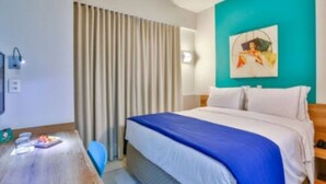 Quarto de hotel com cama, escrivaninha e cadeira, janela com cortinas e um quadro na parede.