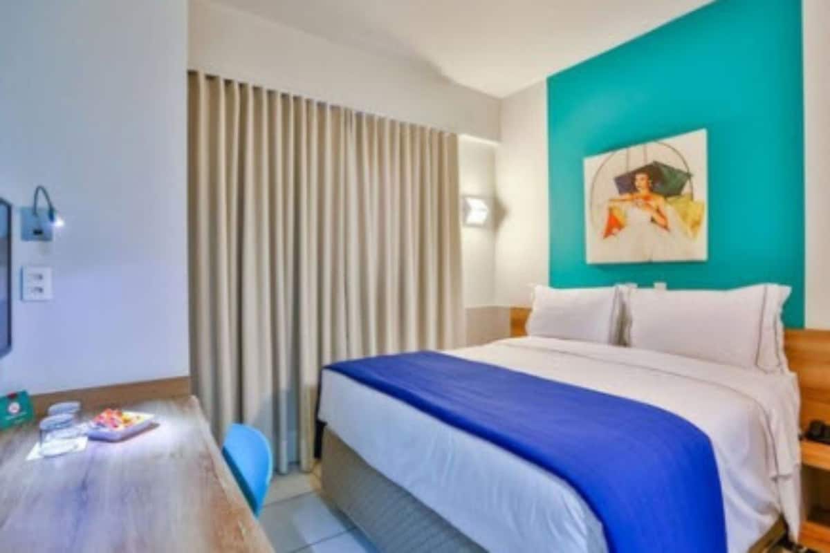 Quarto de hotel com cama, escrivaninha e cadeira, janela com cortinas e um quadro na parede.