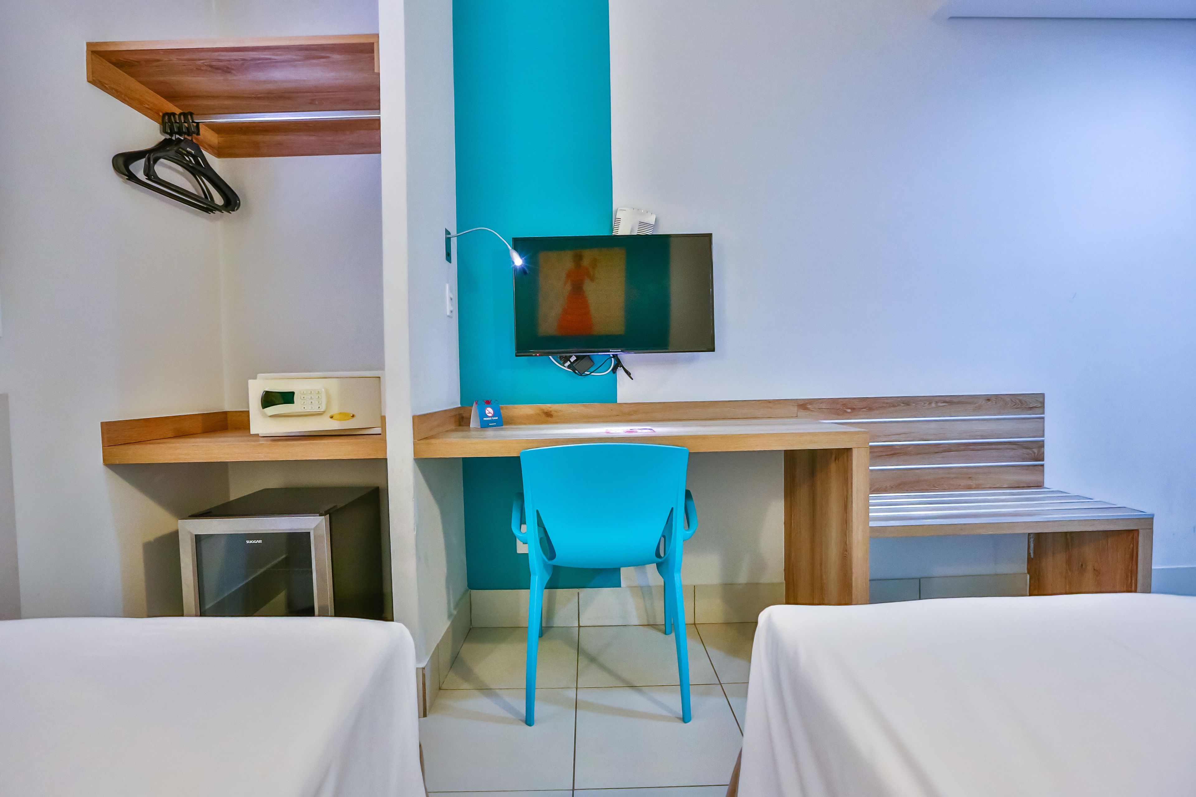 Quarto de hotel com uma escrivaninha de madeira, uma cadeira azul, uma televisão e duas camas.