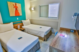 Quarto de hotel com duas camas, uma escrivaninha de madeira, uma janela com persianas, um quadro na parede e uma placa na escrivaninha.