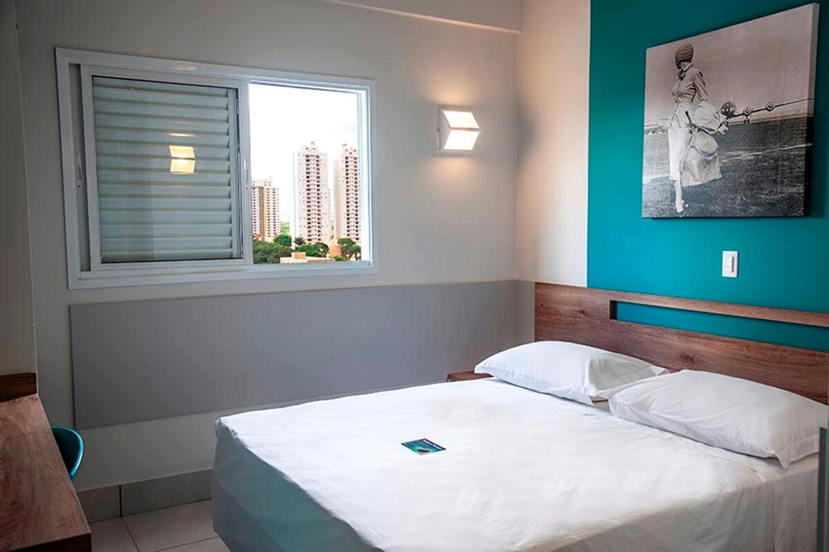 Quarto com cama, janela com persianas, um quadro na parede e vista para prédios.