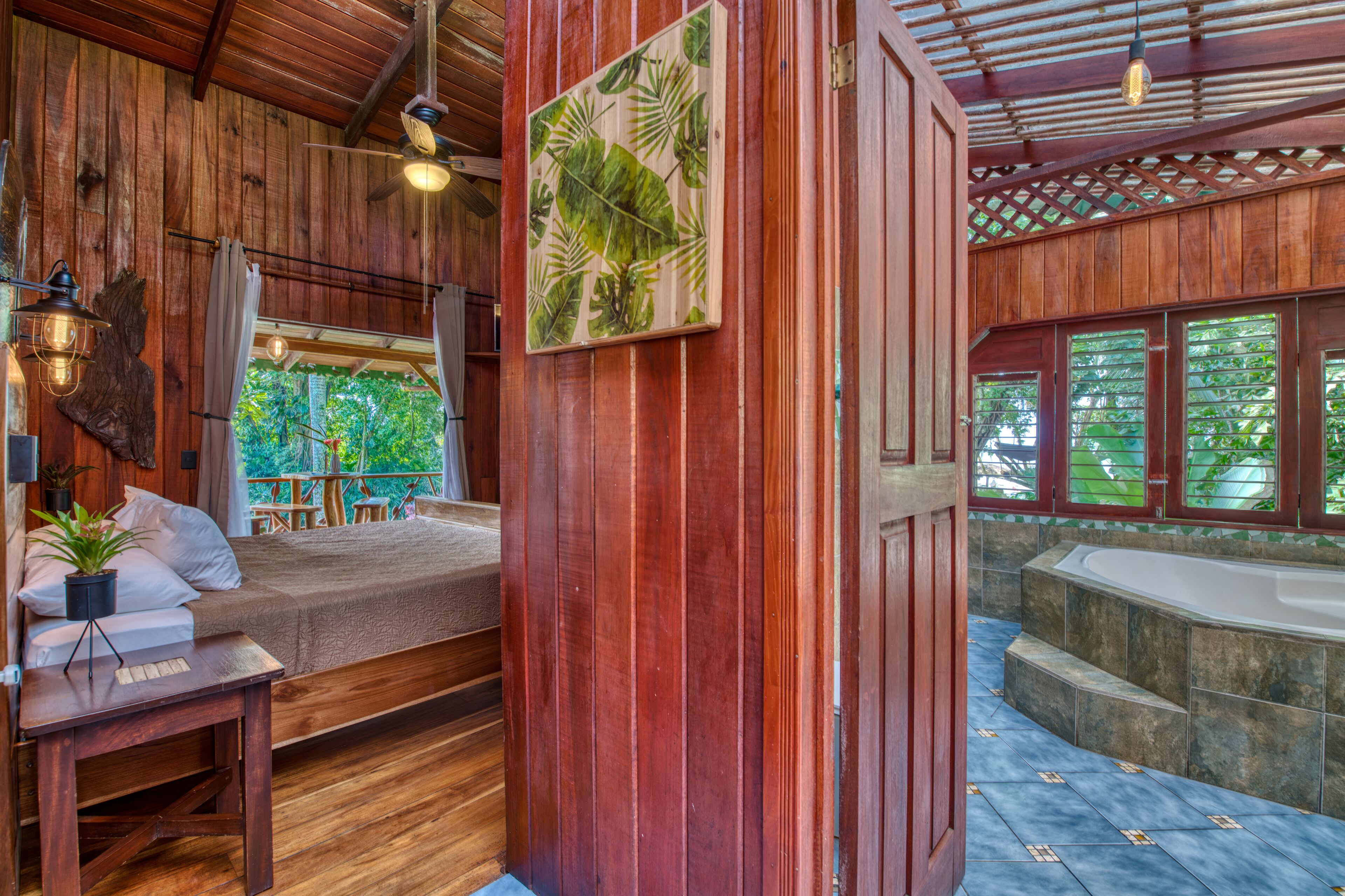 Interior de madeira com uma cama, uma mesinha com uma planta, um quadro e vista para o verde lá fora.