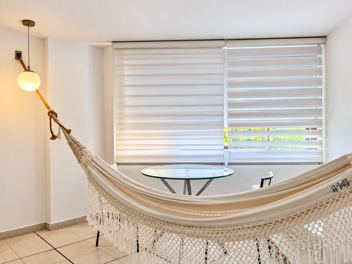 Uma rede em um quarto com uma mesa redonda de vidro, paredes brancas e uma janela com persianas.