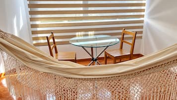 Uma rede com capa de macramé, uma mesa de vidro redonda, duas cadeiras de madeira e uma janela com persianas horizontais.