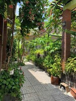 Um caminho estreito ladeado por vegetação exuberante e plantas em vasos, que leva a um edifício com telhado de azulejos.