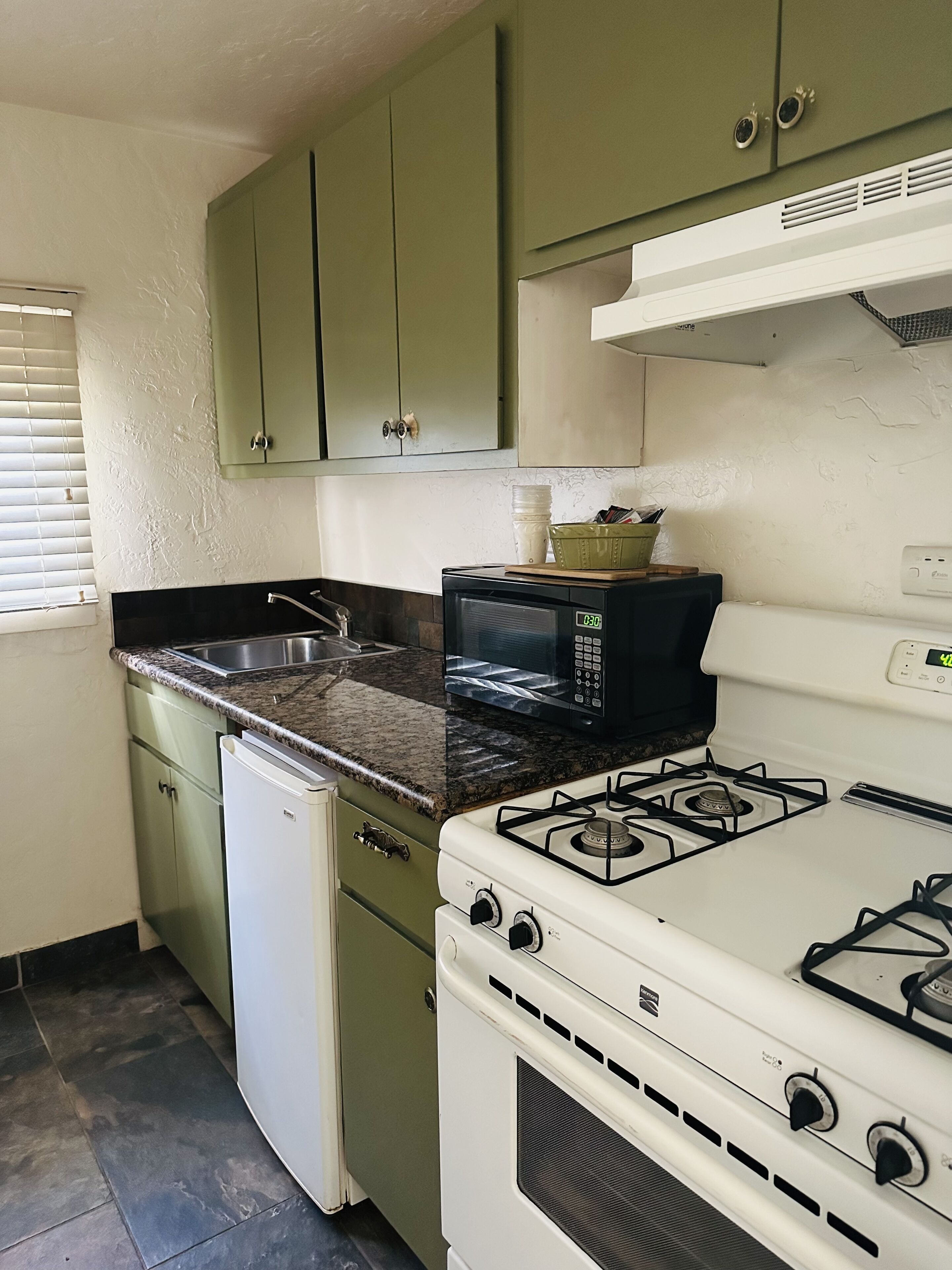 Uma cozinha compacta com armários verdes, um fogão branco, micro-ondas preto e um frigorífico pequeno.