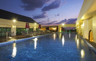Uma grande piscina interior com espreguiçadeiras e guarda-sóis, rodeada por paredes com janelas e luminárias.