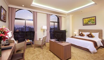 Quarto de hotel com uma cama grande, uma escrivaninha com um computador, uma cadeira e uma televisão. Há grandes janelas com cortinas, um quadro na parede e vista para a paisagem urbana.