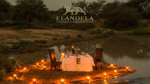 Mesa de jantar posta à beira de um lago, com toalha branca e cadeiras, rodeada por velas.