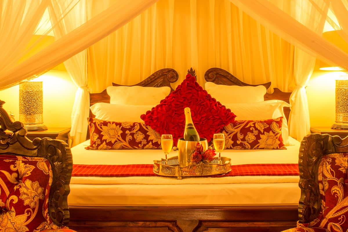 Uma cama luxuosa com dossel, decorada com rosas vermelhas e uma bandeja com taças de champanhe e uma garrafa.