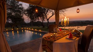 Área de refeições ao ar livre com vista para piscina, mesa iluminada por velas e um pôr do sol ao fundo.