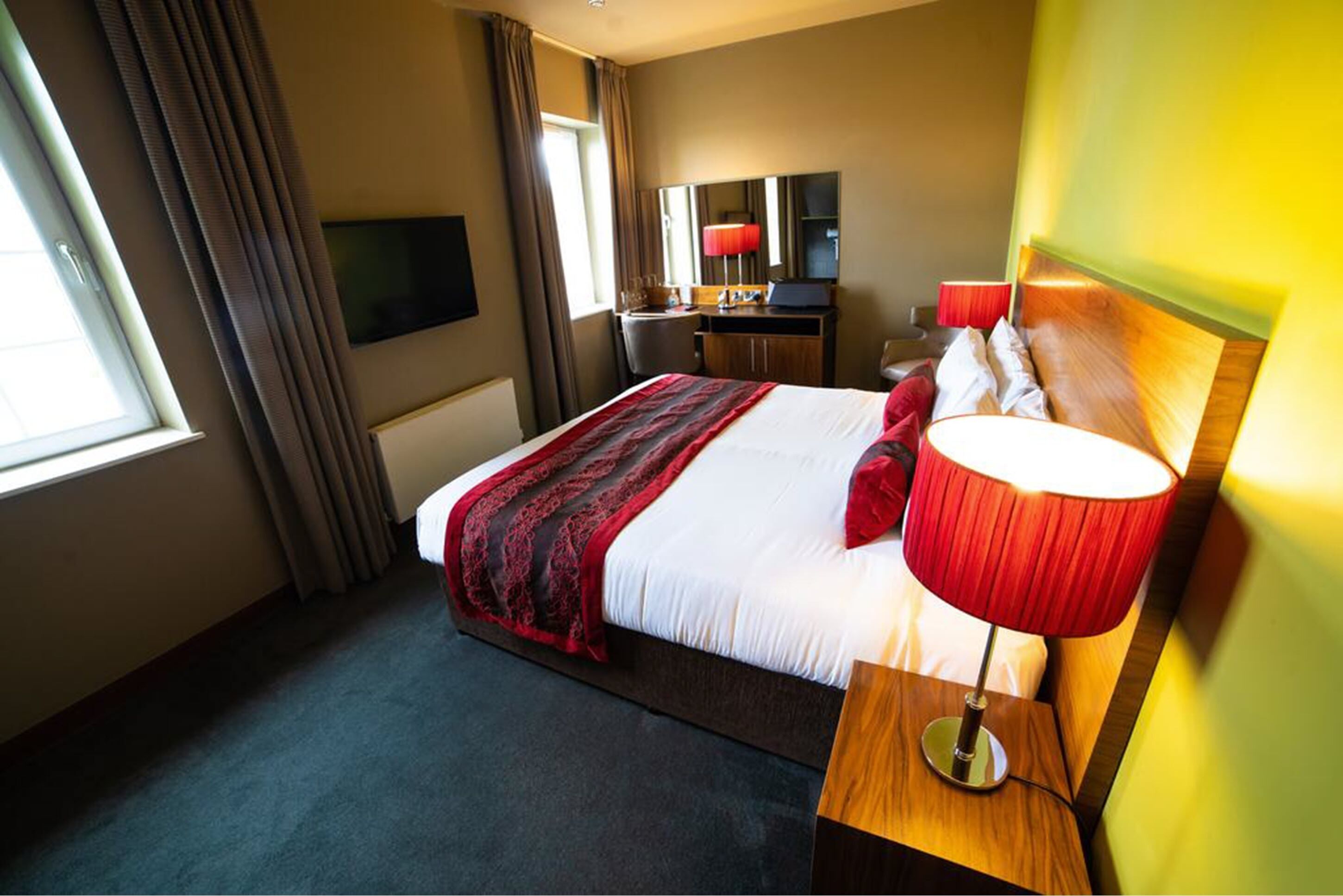 Quarto de hotel com cama, escrivaninha, cadeira, televisão e janela com cortinas.