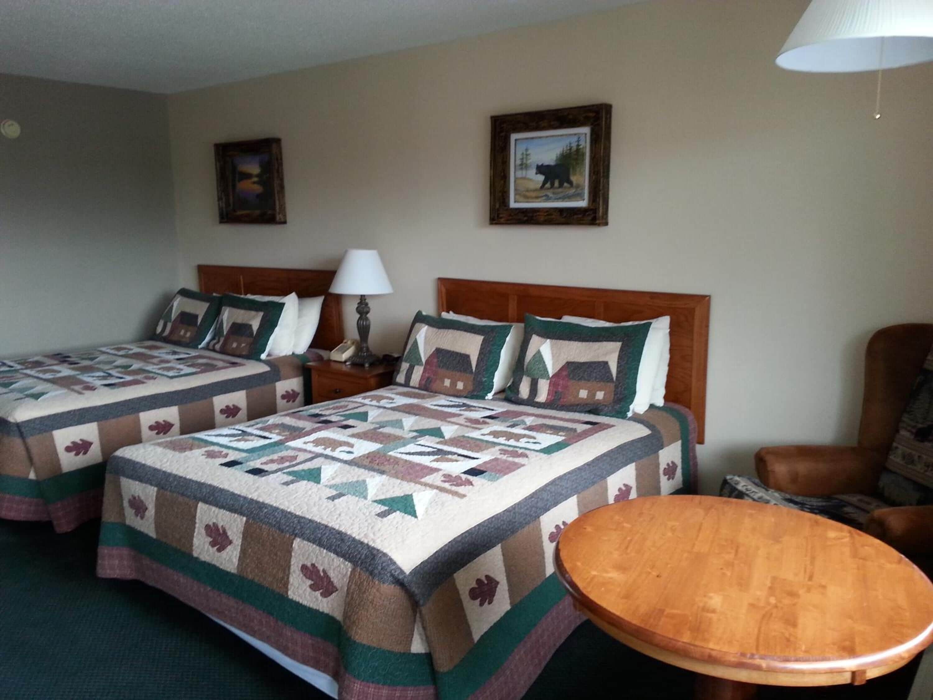 Quarto de hotel com duas camas, cabeceira de madeira, mesa de cabeceira com abajur, mesa redonda de madeira e duas quadros decorativos na parede.