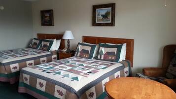 Quarto de hotel com duas camas, cabeceira de madeira, mesa de cabeceira com abajur, mesa redonda de madeira e duas quadros decorativos na parede.