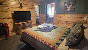Um quarto acolhedor com teto e paredes de madeira, uma cama com colcha estampada, uma televisão sobre um suporte de madeira e uma porta que dá para outro quarto.