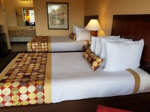 Quarto de hotel com duas camas, cada uma com roupa de cama branca e colchas estampadas. Há uma cabeceira de madeira, um abajur e um espelho na parede.