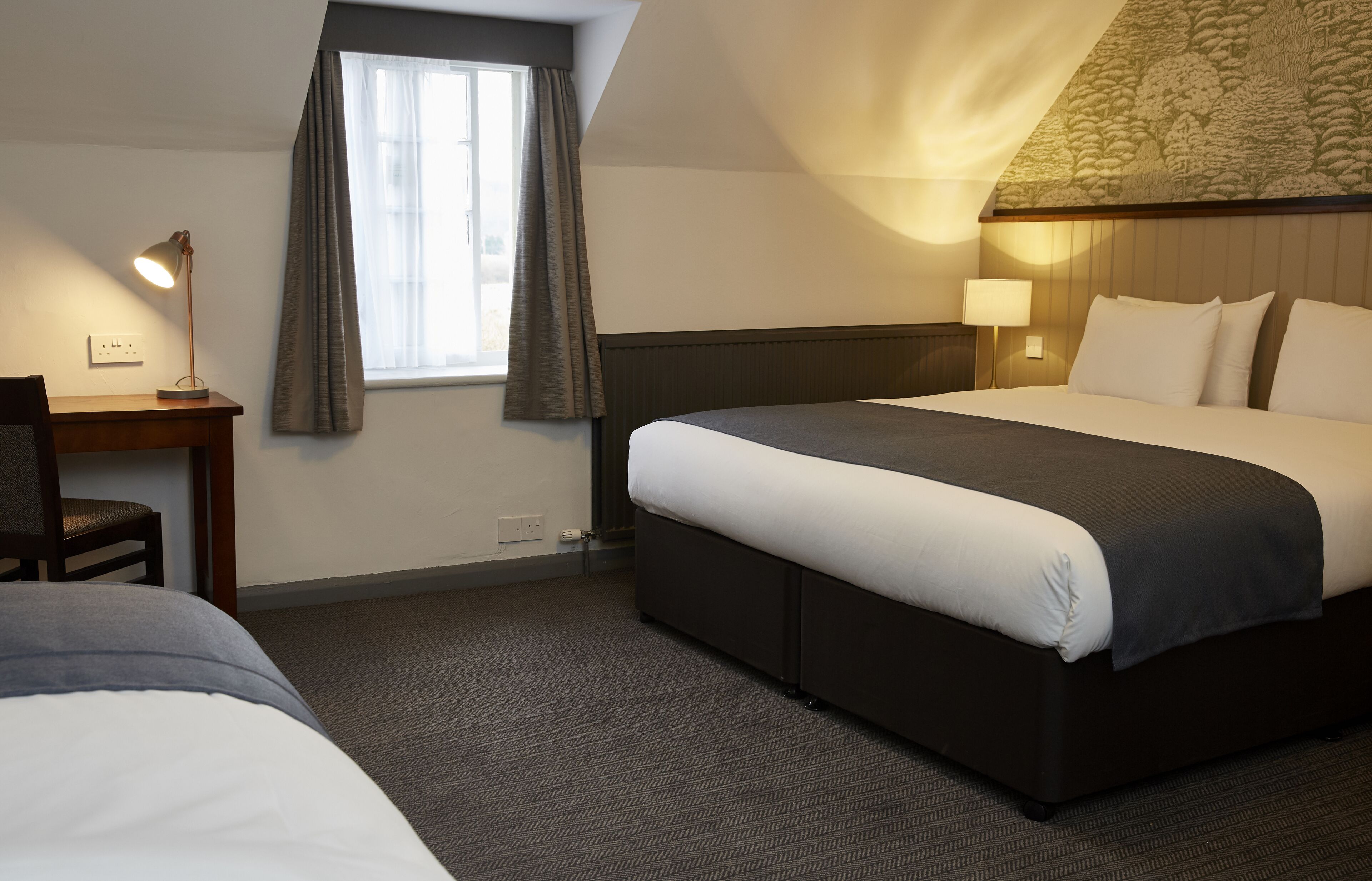 Quarto de hotel com uma cama grande, uma mesa com abajur, uma janela com cortinas e uma parede estampada.