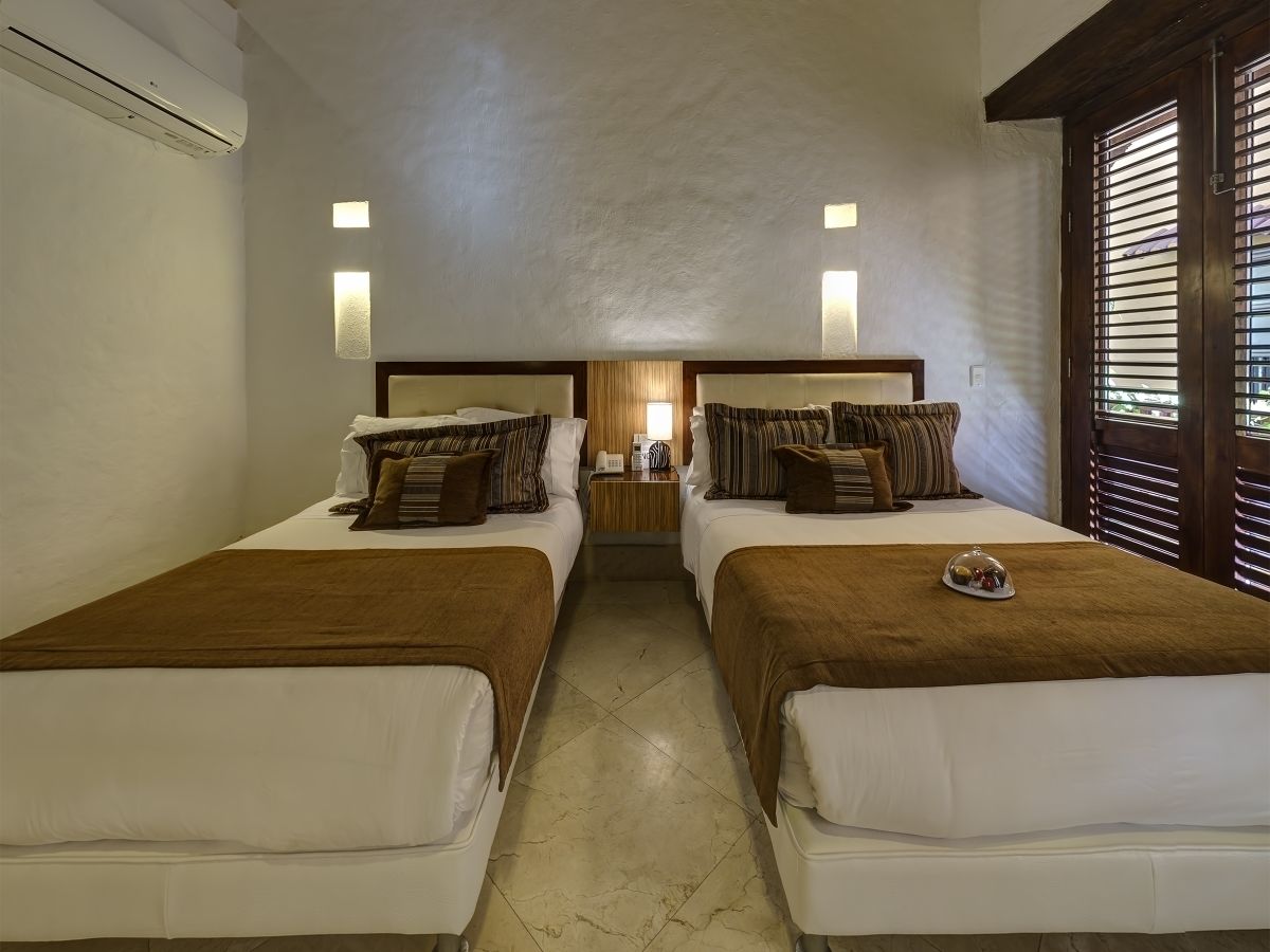 Quarto de hotel com duas camas de solteiro, cabeceira de madeira, mesa de cabeceira com abajur e janela com persianas.