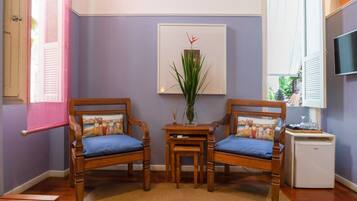 Quarto com duas cadeiras de madeira com almofadas azuis, uma pequena mesa de madeira, uma obra de arte emoldurada na parede, um refrigerador pequeno e uma janela com persianas.