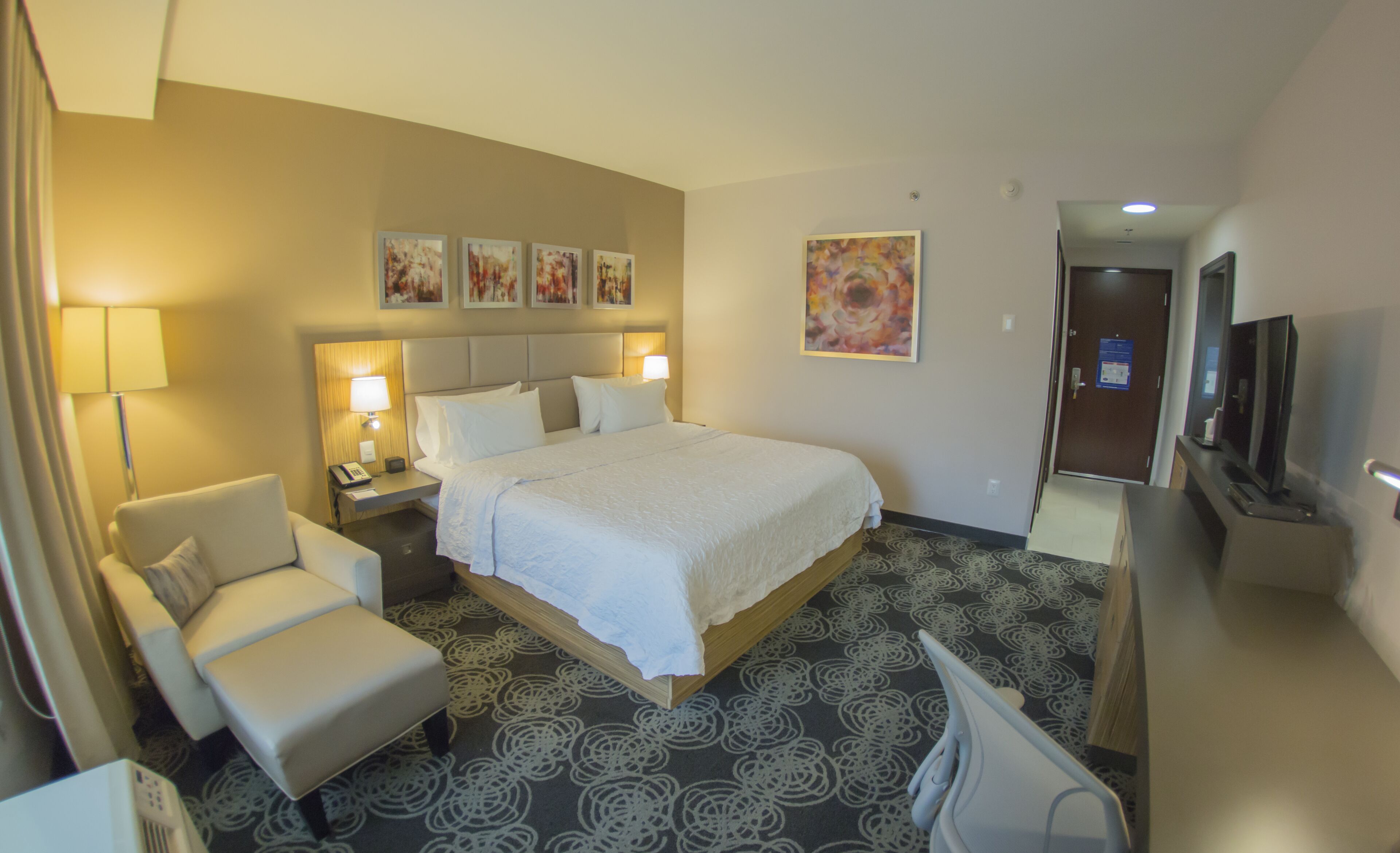 Quarto de hotel com uma cama grande, escrivaninha, cadeira, televisão e obras de arte nas paredes.