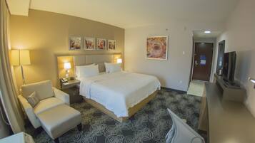 Quarto de hotel com uma cama grande, escrivaninha, cadeira, televisão e obras de arte nas paredes.