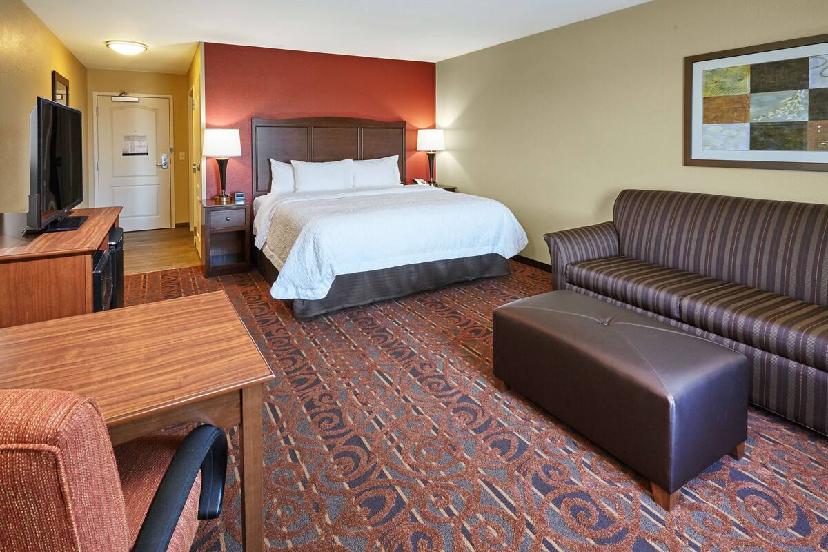 Quarto de hotel com uma cama, um sofá, uma mesa de escritório com cadeira, uma televisão e um abajur.