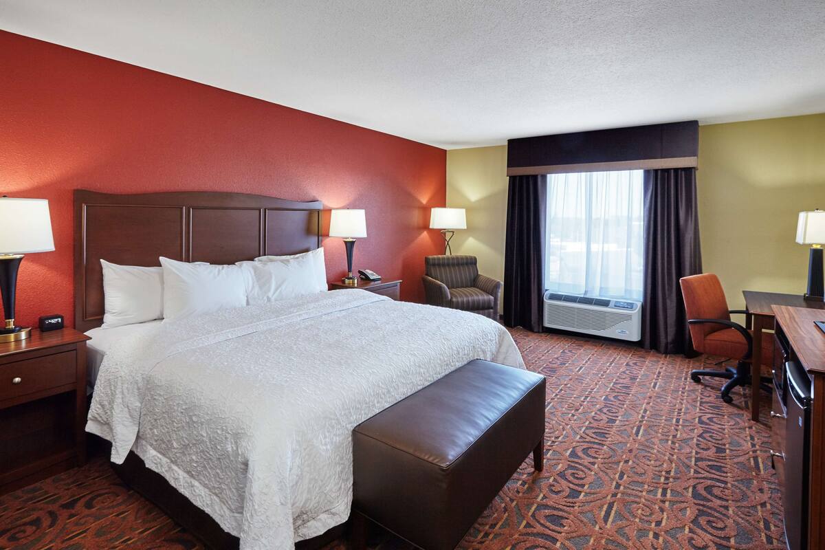 Quarto de hotel com uma cama grande, duas mesas de cabeceira com luminárias, uma cadeira, uma escrivaninha e uma janela com cortinas.
