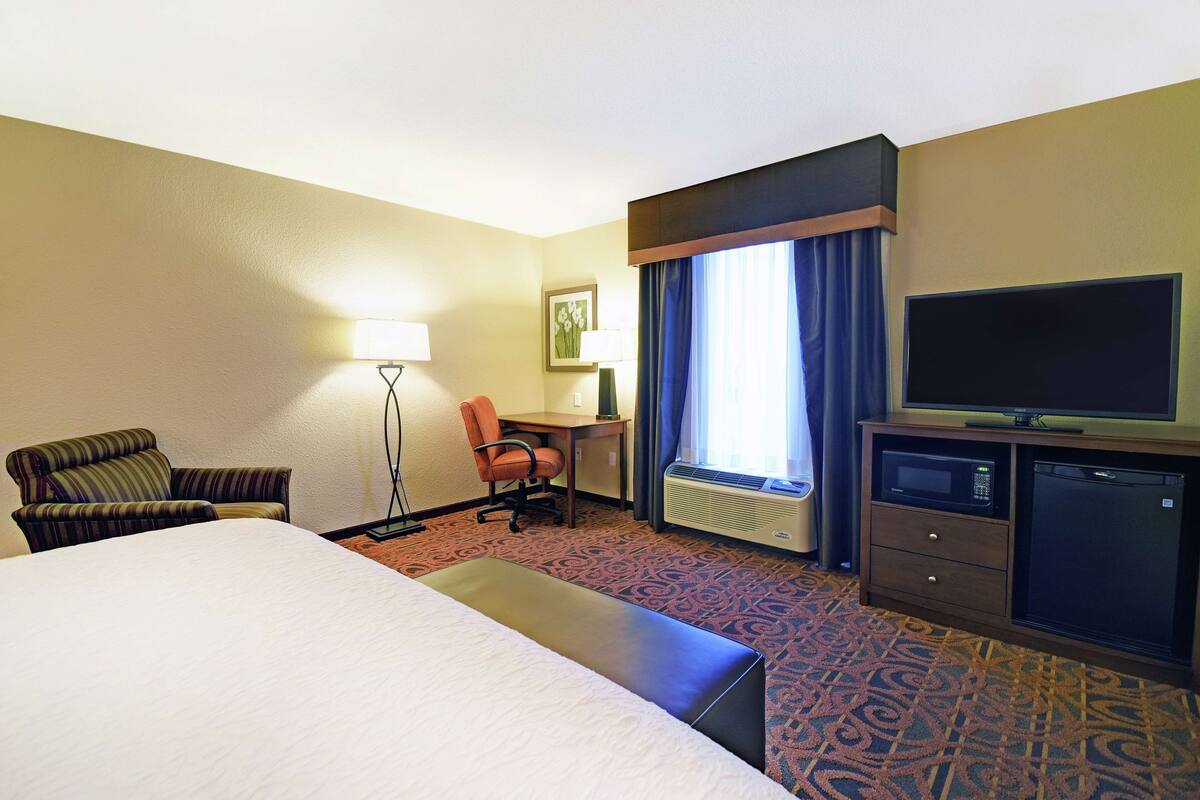 Quarto de hotel com uma cama grande, uma escrivaninha com cadeira, televisão e minibar.